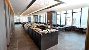 Um saguão de hotel moderno com um longo balcão de buffet, área de estar e amplas janelas com vista para a paisagem urbana.