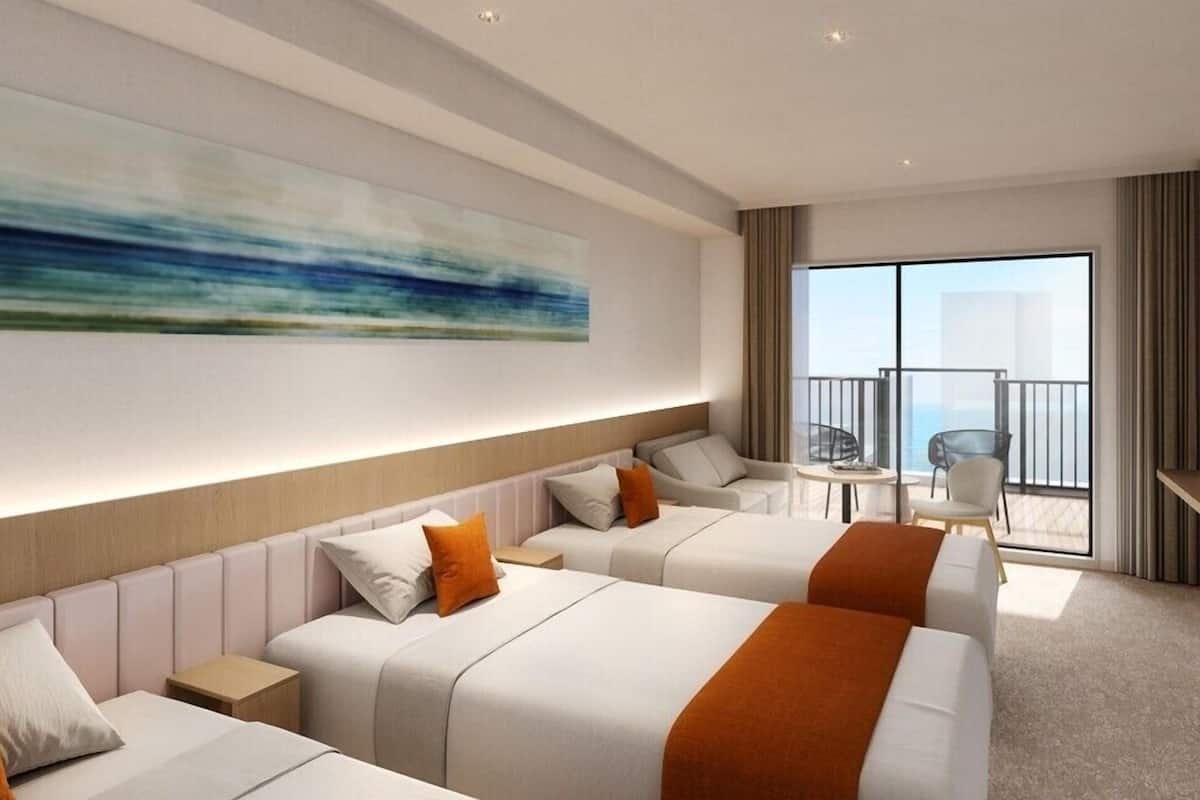 Quarto de hotel com três camas, uma grande pintura abstrata, varanda com vista e uma pequena mesa com cadeiras.