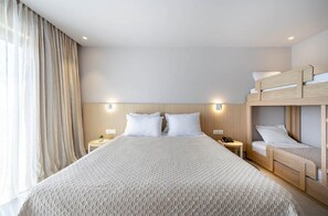 Quarto de hotel com uma cama grande, duas mesinhas de cabeceira, uma cama beliche e uma janela com cortinas.