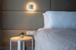 Quarto de hotel com uma cama, uma mesa de cabeceira com uma planta, uma luminária na parede e uma tomada.