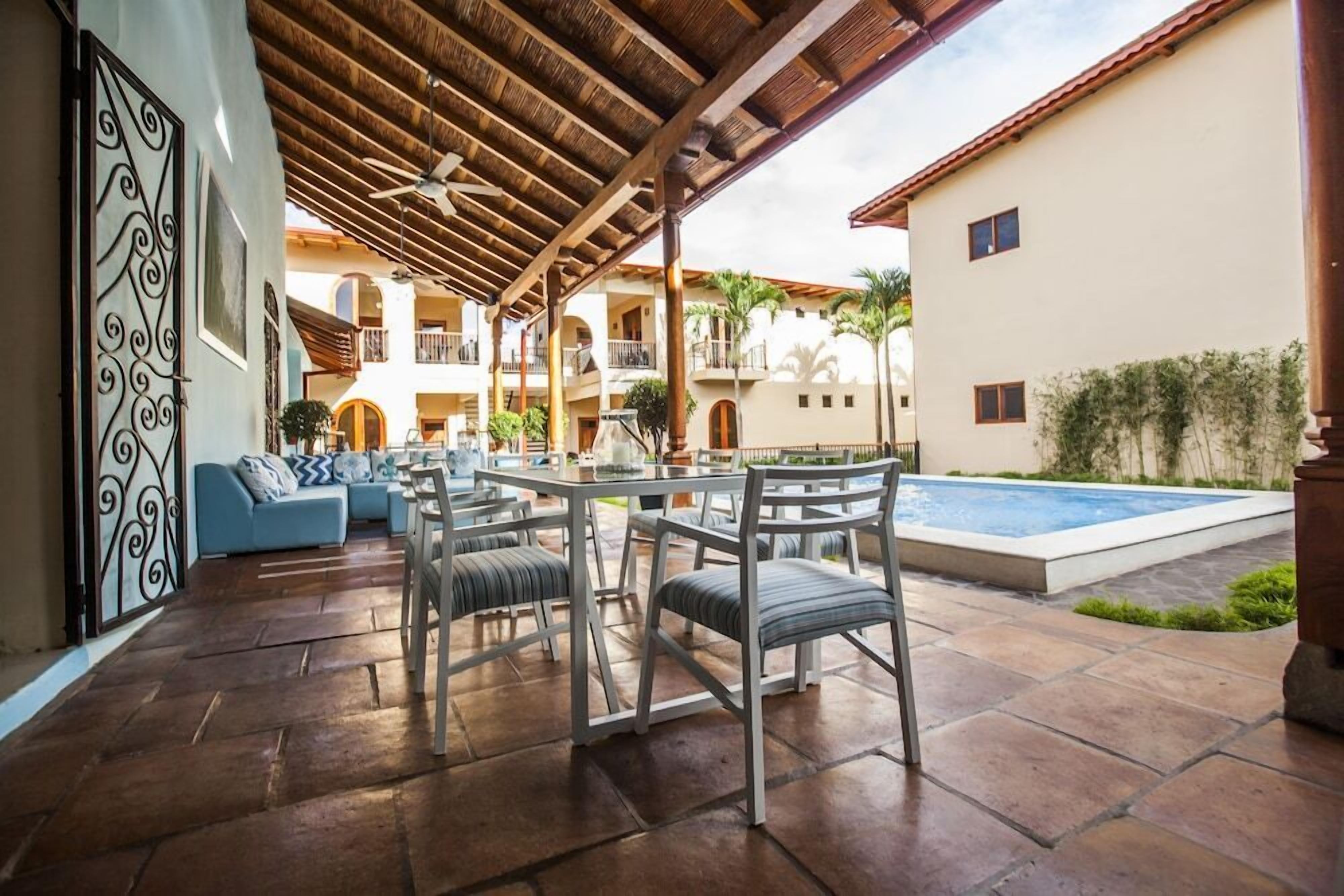 Uma área de estar coberta ao ar livre com mesas e cadeiras, piscina e um edifício com varandas.