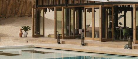 Uma área de piscina com espreguiçadeiras, um espaço de estar coberto e vista para a praia.