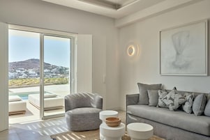 Uma sala de estar moderna com um sofá cinzento, uma poltrona cinzenta e uma mesa de centro. Há uma janela grande com vista para uma paisagem montanhosa.