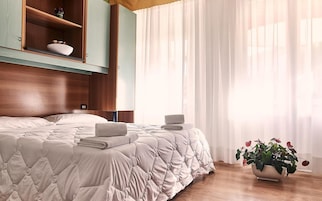 Cama bem arrumada com cobertura acolchoada, mesa de cabeceira com toalha dobrada, prateleira de madeira com objetos decorativos e planta em vaso.