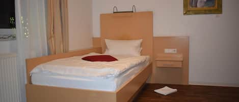 Uma cama de solteiro com estrutura de madeira, uma mesinha de cabeceira e uma janela com cortinas.