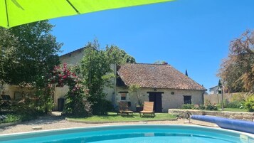 Uma área de piscina com céu azul límpido, um edifício de pedra com telhado telhado e um guarda-sol verde.
