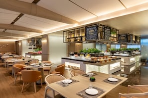Interior moderno de restaurante com várias mesas para refeições, uma área central de buffet e diversos alimentos expostos.
