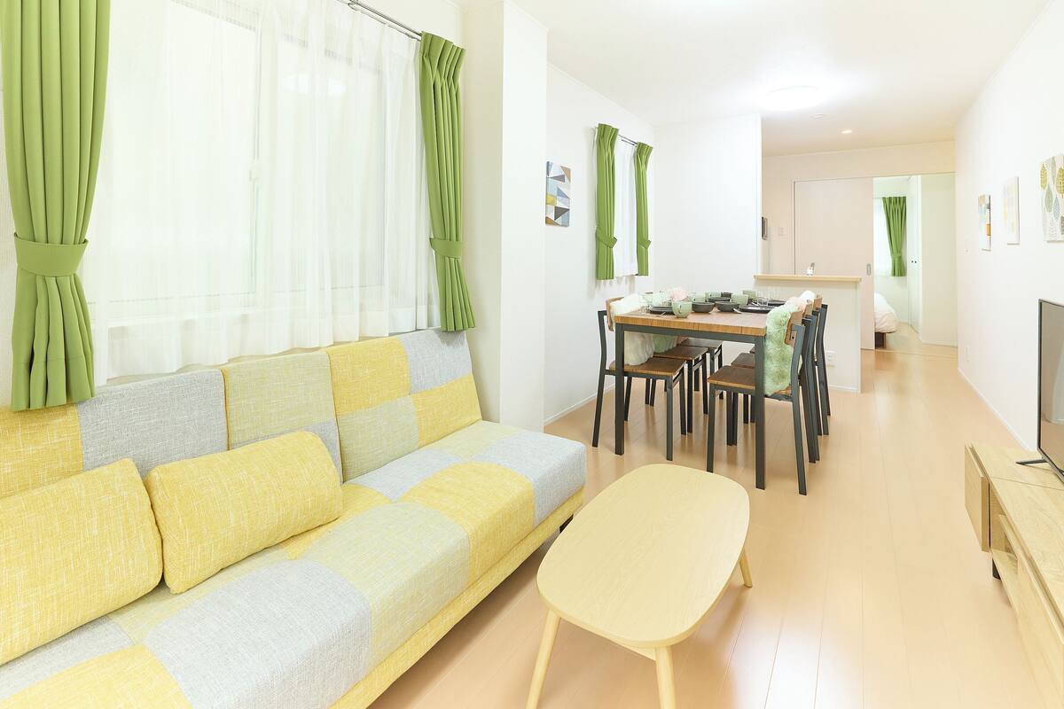 Uma sala de estar com sofá, mesa de jantar com cadeiras, televisão e uma área de cozinha ao fundo.