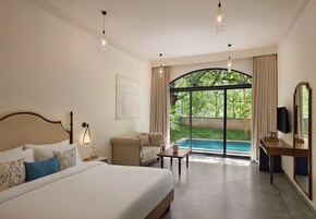 Quarto de hotel com cama, sofá, mesa, televisão e vista para uma piscina.
