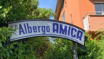 Placa de 'Albergo AMICA', com uma cerca de metal e vegetação ao fundo.