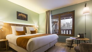 Quarto de hotel com uma cama grande, uma mesa redonda pequena, uma cadeira, uma janela com vista para edifícios e uma luminária.