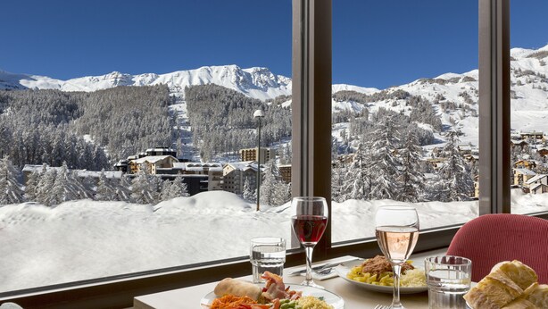 Mesa de jantar com comida e copos, com vista para uma paisagem nevada de montanha através de uma janela.