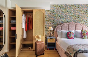 Quarto de hotel com guarda-roupa, mala, cama com almofadas, mesa de cabeceira e parede com padrão floral.