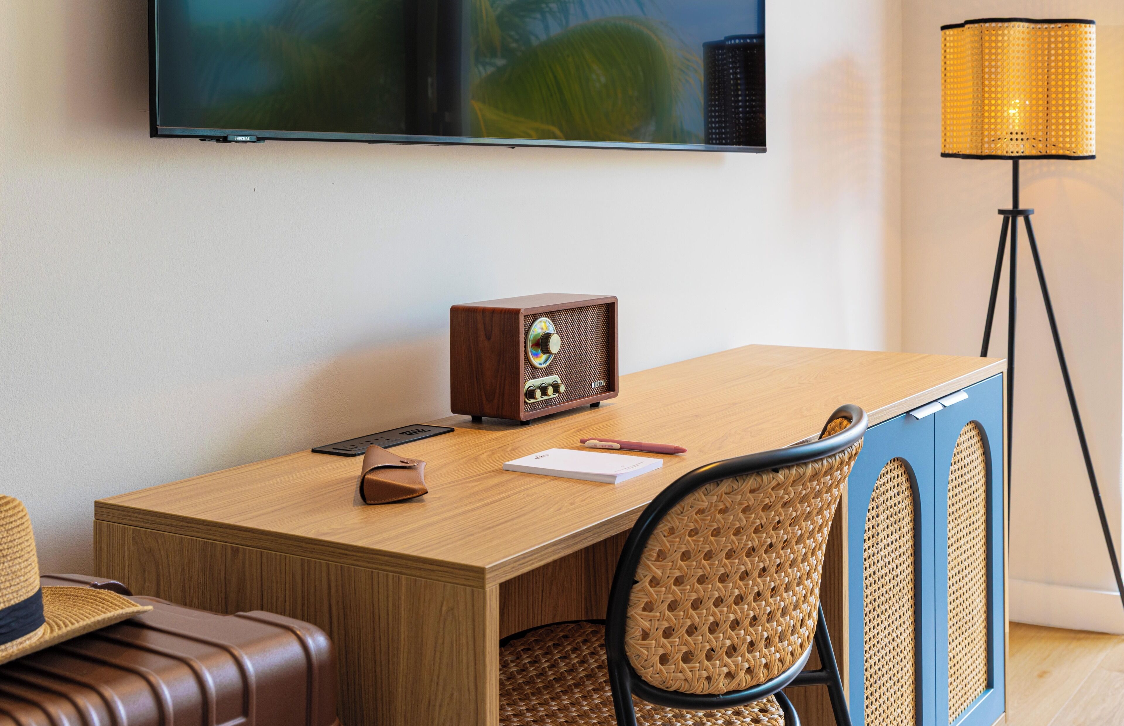 Quarto de hotel com uma escrivaninha de madeira, um rádio antigo, televisão fixada na parede, abajur e uma cadeira de vime.