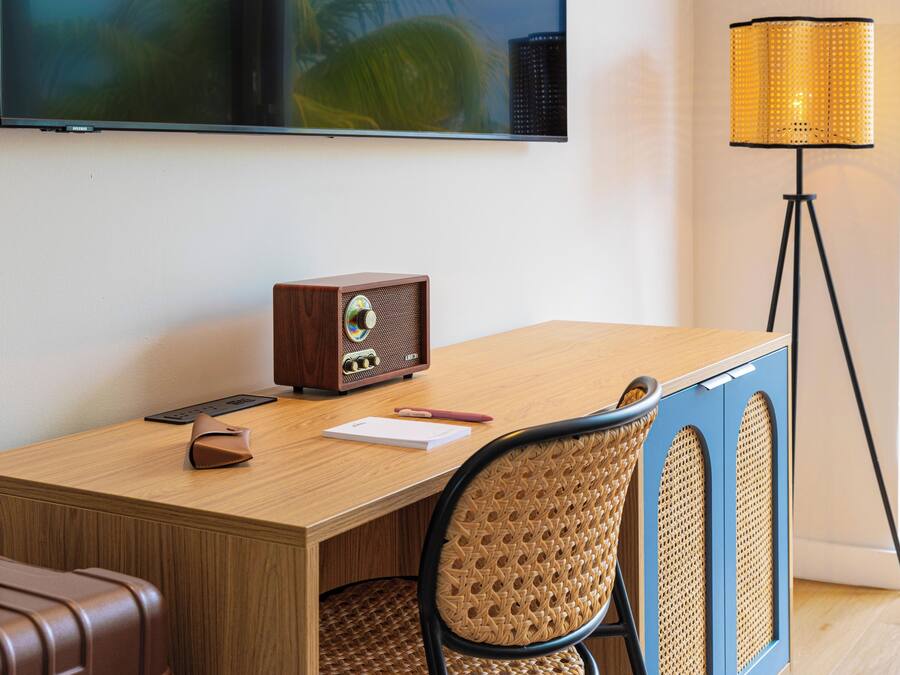 Quarto de hotel com uma escrivaninha de madeira, um rádio antigo, televisão fixada na parede, abajur e uma cadeira de vime.