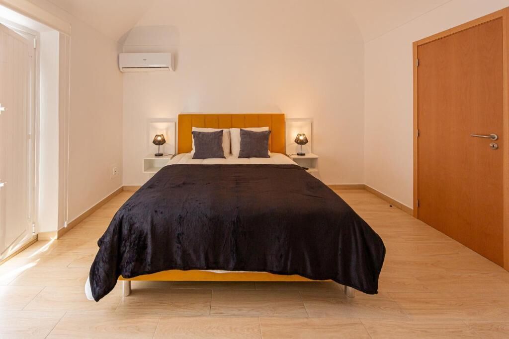 Quarto com cama, luminárias de cabeceira, porta de madeira e ar condicionado.