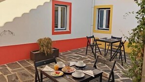 Uma área de refeições ao ar livre com piso de pedra, mesas e cadeiras de madeira e duas janelas com molduras vermelhas e amarelas.