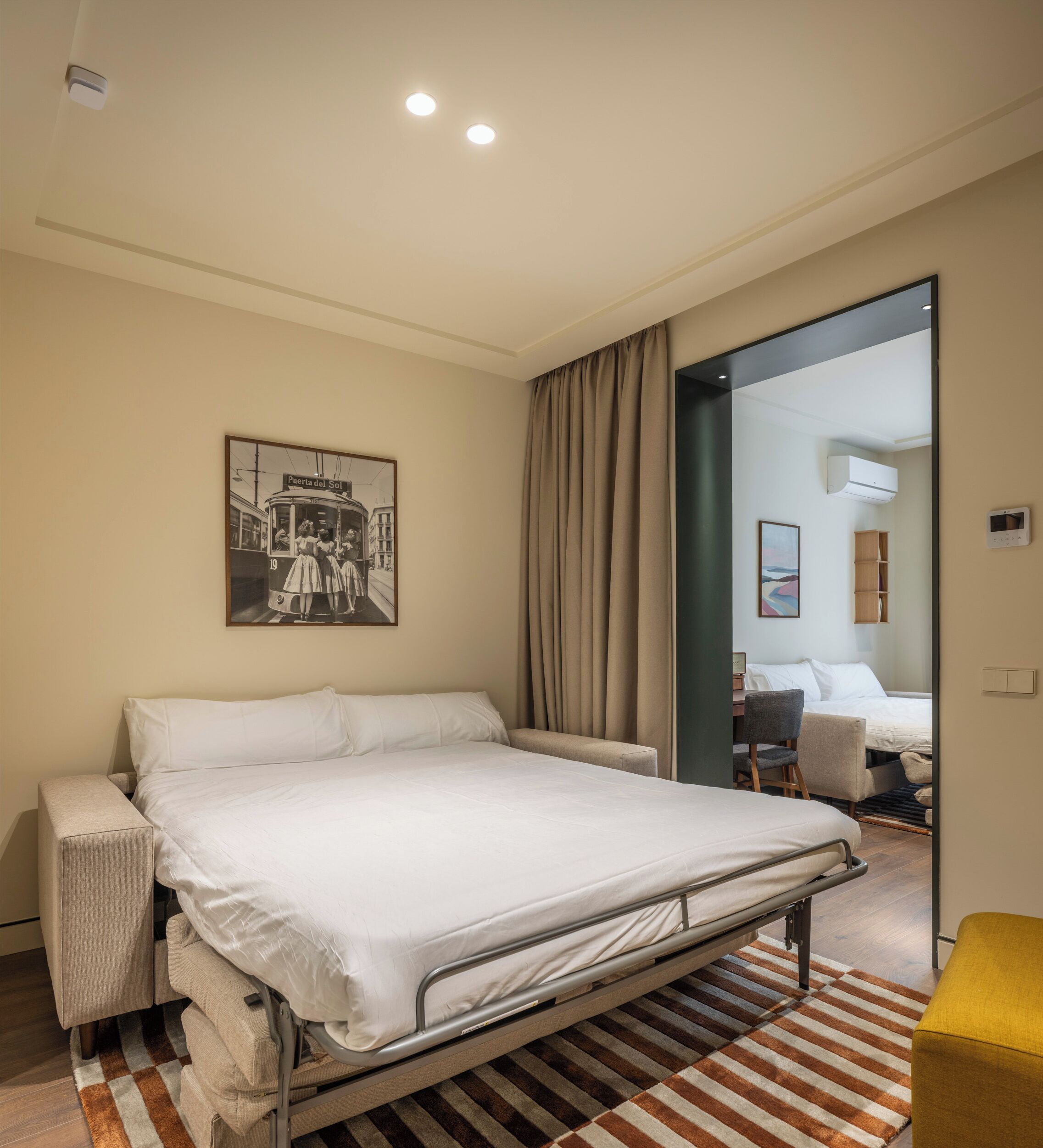 Quarto de hotel com cama, uma cadeira, escrivaninha, espelho e um quadro na parede.