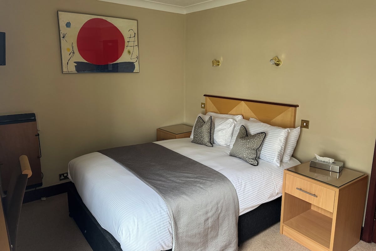 Quarto de hotel com uma cama grande, duas mesas de cabeceira, um criado-mudo, uma obra de arte na parede e uma cabeceira de madeira.