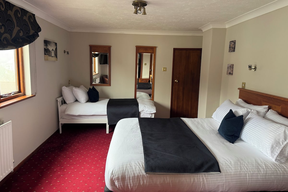 Quarto de hotel com duas camas, um tapete vermelho, um ventilador de teto e uma janela com cortinas.
