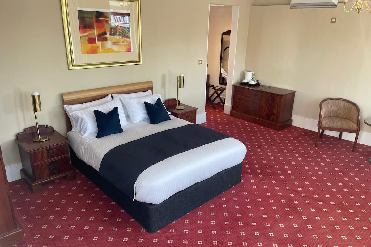 Quarto de hotel com cama grande, mesa de cabeceira, cômoda, cadeira e um quadro na parede.