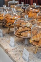 Vitrine de pastelaria com diversos doces sob cúpulas de vidro, identificados com nomes como 'ALMOND CROISSANT'.