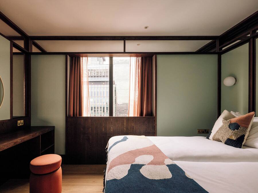 Quarto de hotel com uma cama grande, um banco de madeira, um espelho redondo e uma janela com cortinas.
