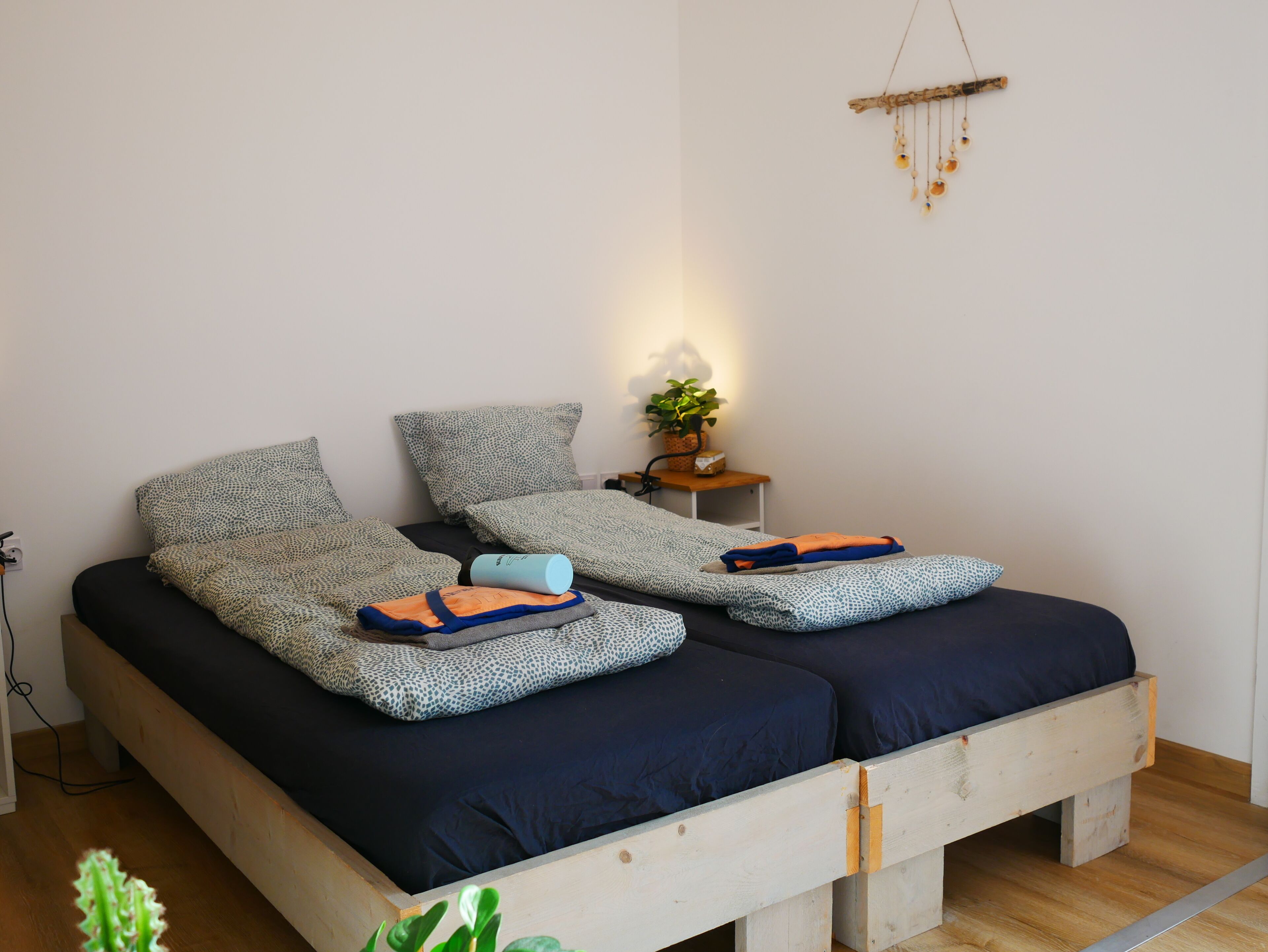 Uma cama com duas almofadas, uma mesa de cabeceira com abajur e um adorno pendurado.