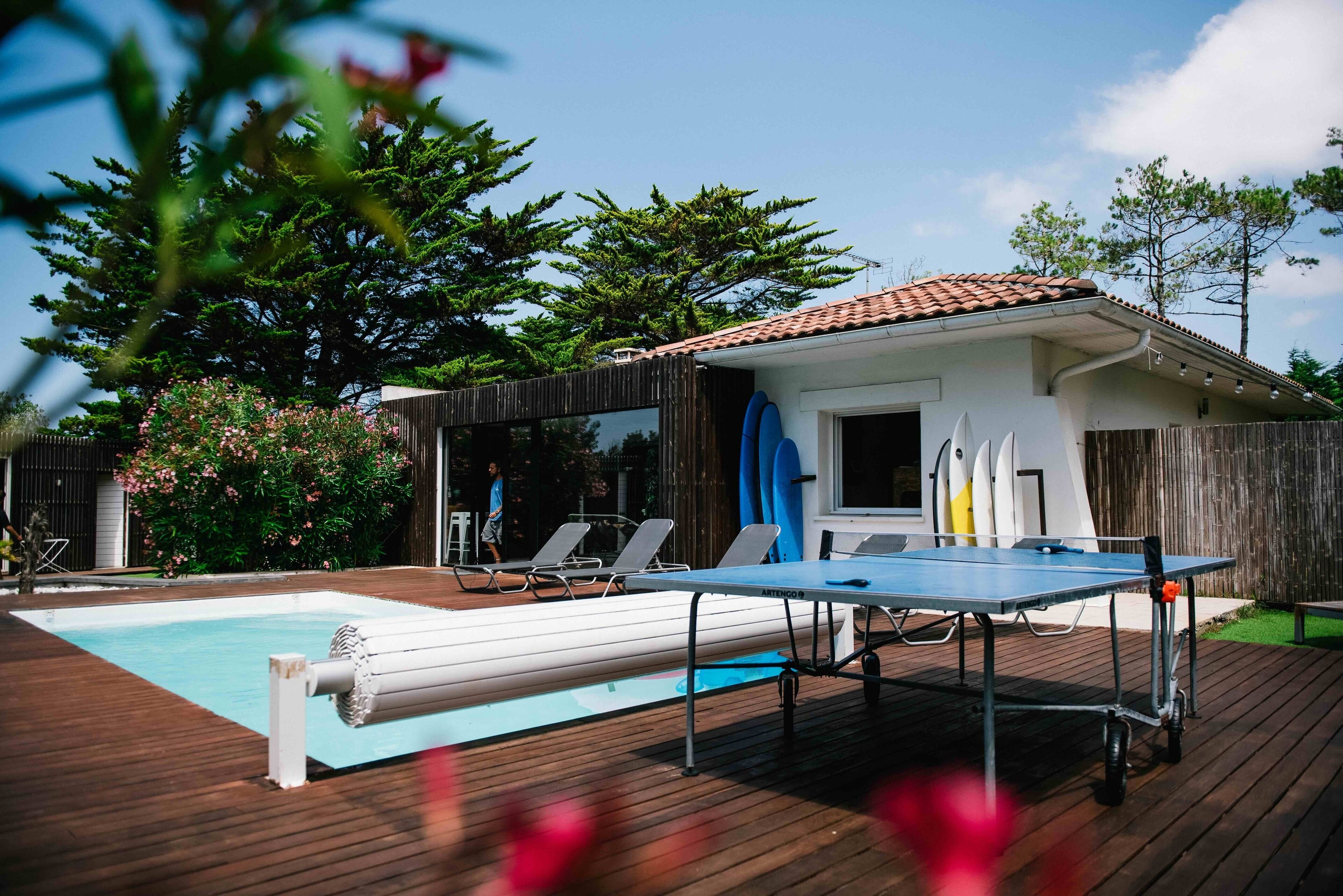 Uma área à beira da piscina com mesa, espreguiçadeiras e um pequeno edifício com telhado em azulejo.