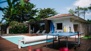 Uma área à beira da piscina com mesa, espreguiçadeiras e um pequeno edifício com telhado em azulejo.