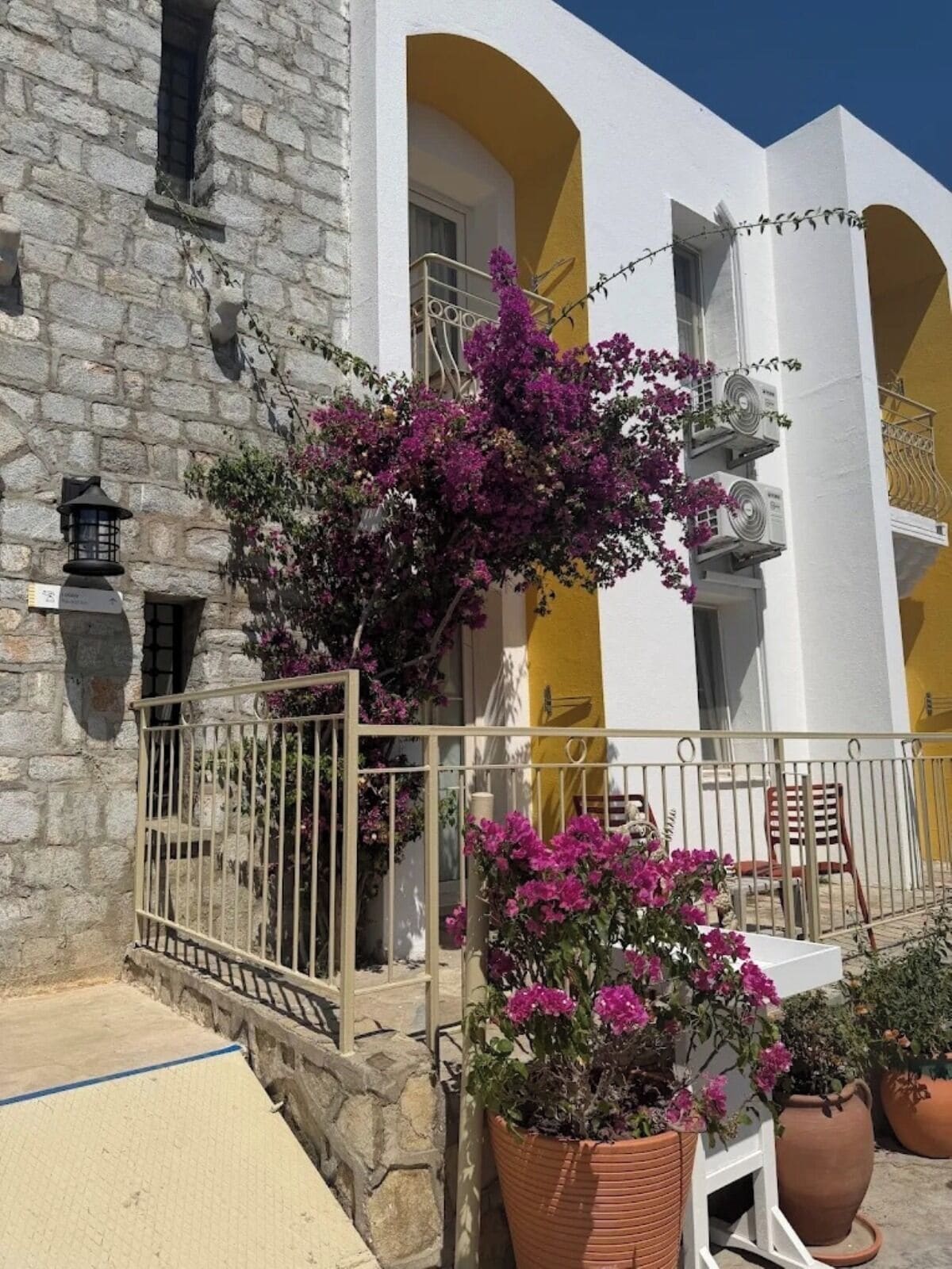 Um edifício com paredes de pedra, uma varanda com grade de metal e vasos com flores roxas.