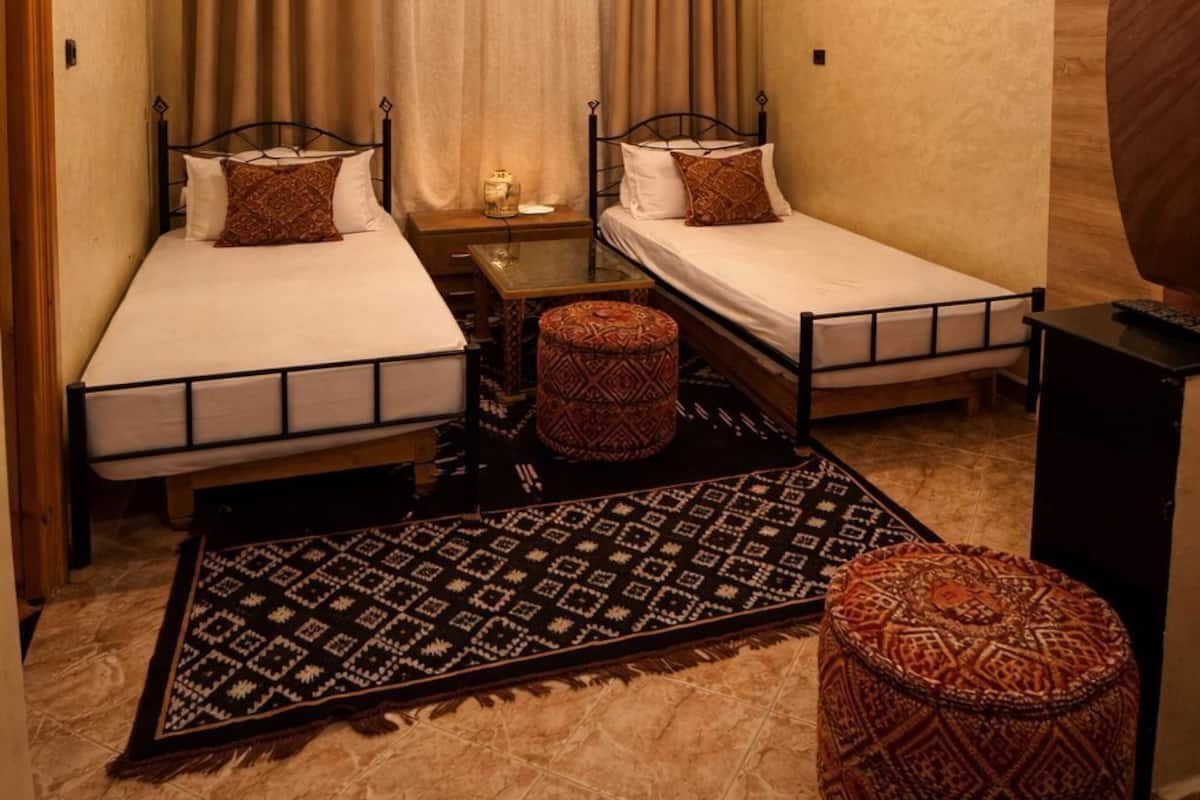 Quarto compacto com duas camas de solteiro, uma mesinha, dois pufes e um lustre no teto.
