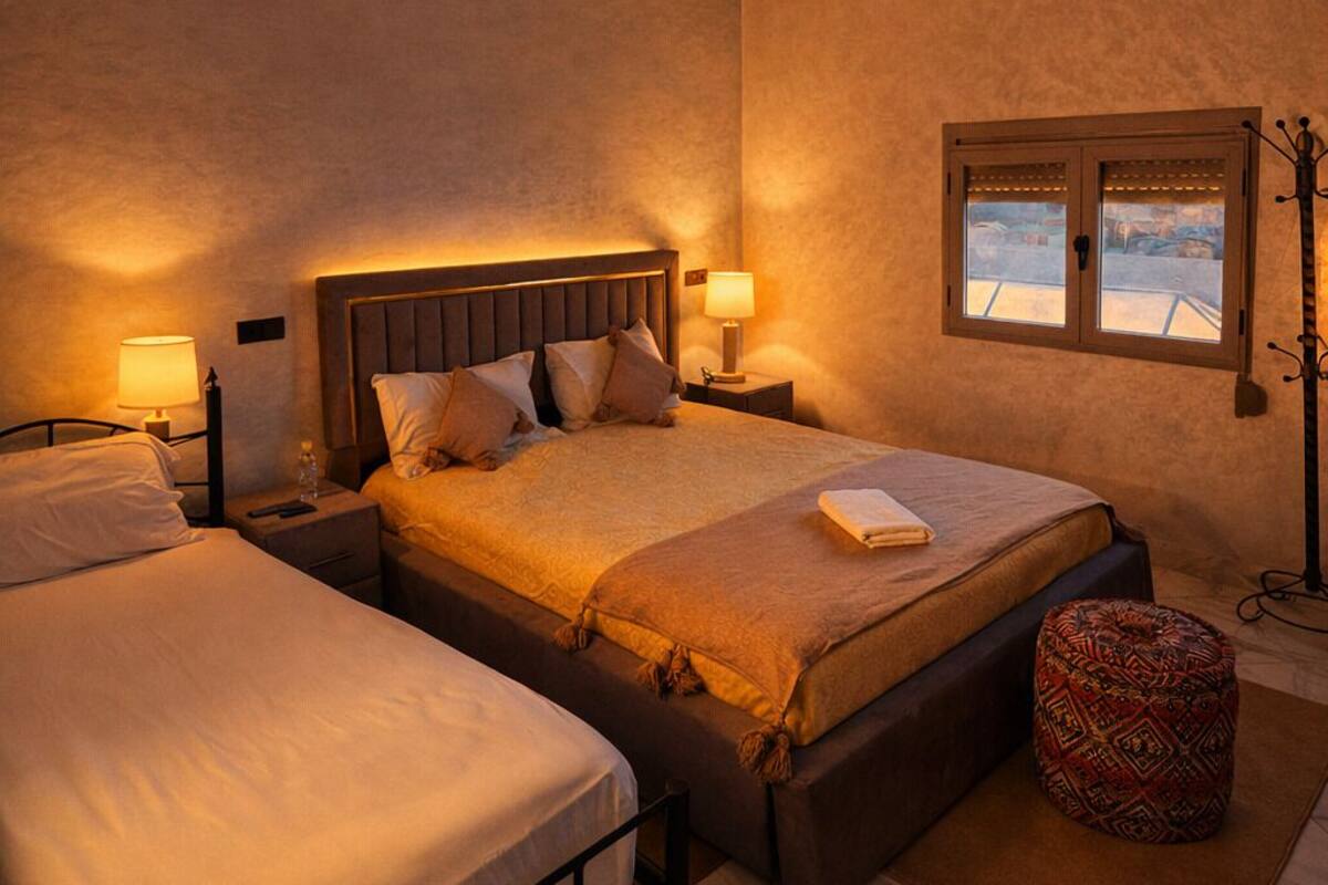 Quarto de hotel com duas camas, janela com vista e um banquinho estampado vermelho.