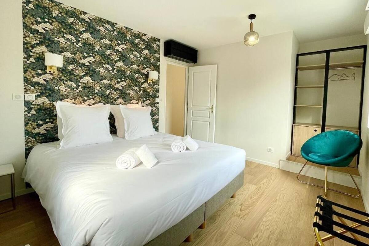 Uma cama bem arrumada com lençóis brancos, parede com padrão floral, uma cadeira moderna e um guarda-roupa com espelho.