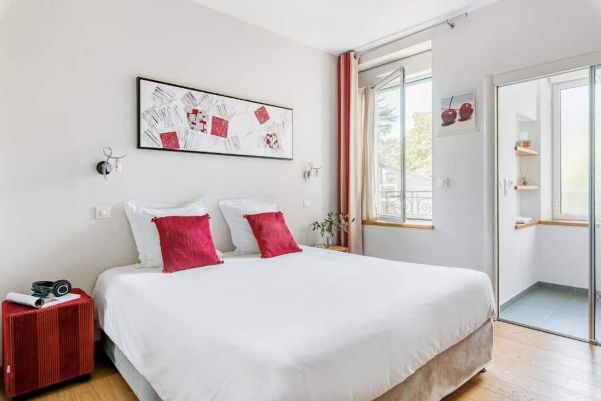 Quarto moderno com uma cama grande, um travesseiro decorativo vermelho, uma obra de arte abstrata emoldurada e uma porta de correr de vidro para uma varanda.