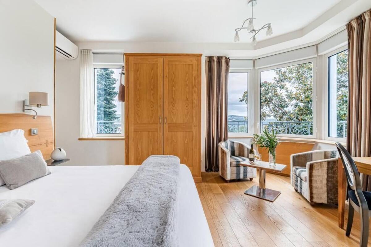 Quarto com uma cama grande, guarda-roupa de madeira, área de estar com mesa e cadeiras, e janelas amplas com vista para o exterior.