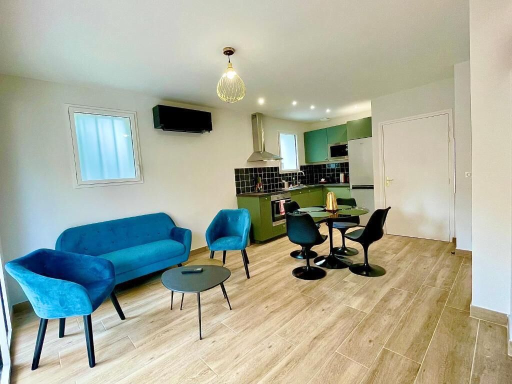 Uma sala de estar moderna com um sofá azul, uma mesa de centro e cadeiras pretas. Uma cozinha com armários verdes e um micro-ondas é visível ao fundo.