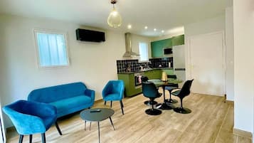 Uma sala de estar moderna com um sofá azul, uma mesa de centro e cadeiras pretas. Uma cozinha com armários verdes e um micro-ondas é visível ao fundo.