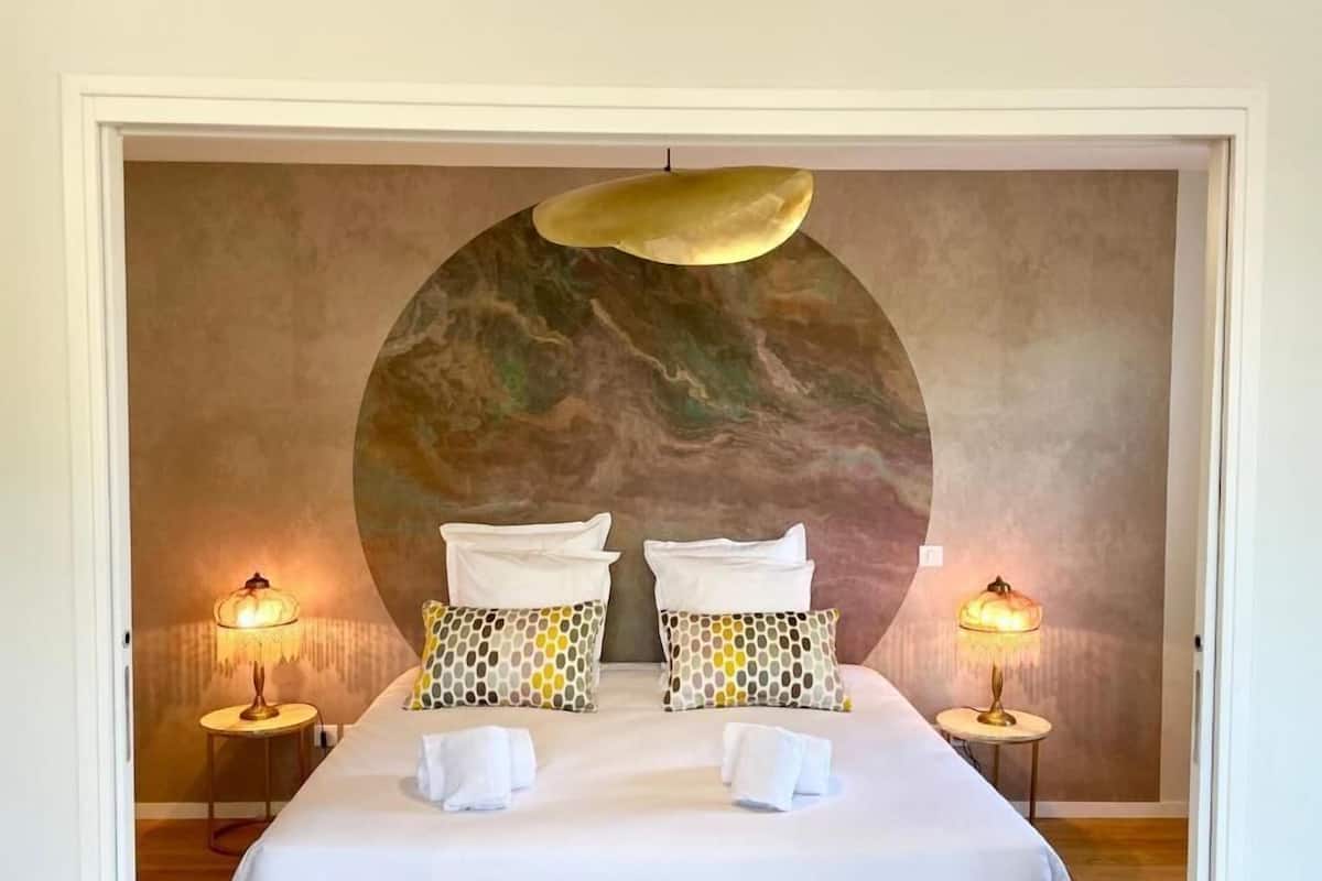 Cama bem arrumada com lençóis brancos e almofadas estampadas, duas luminárias de cabeceira e um quadro abstrato grande na parede.