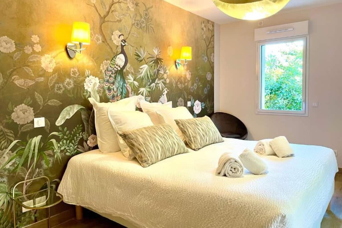 Quarto com cama, almofadas, mesa de cabeceira, luminária e janela com vista para vegetação.