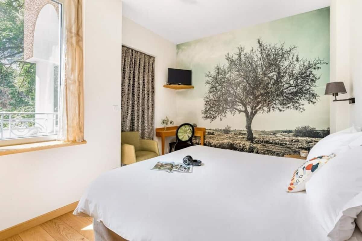 Quarto com cama, relógio, televisão numa estante e janela com cortinas.