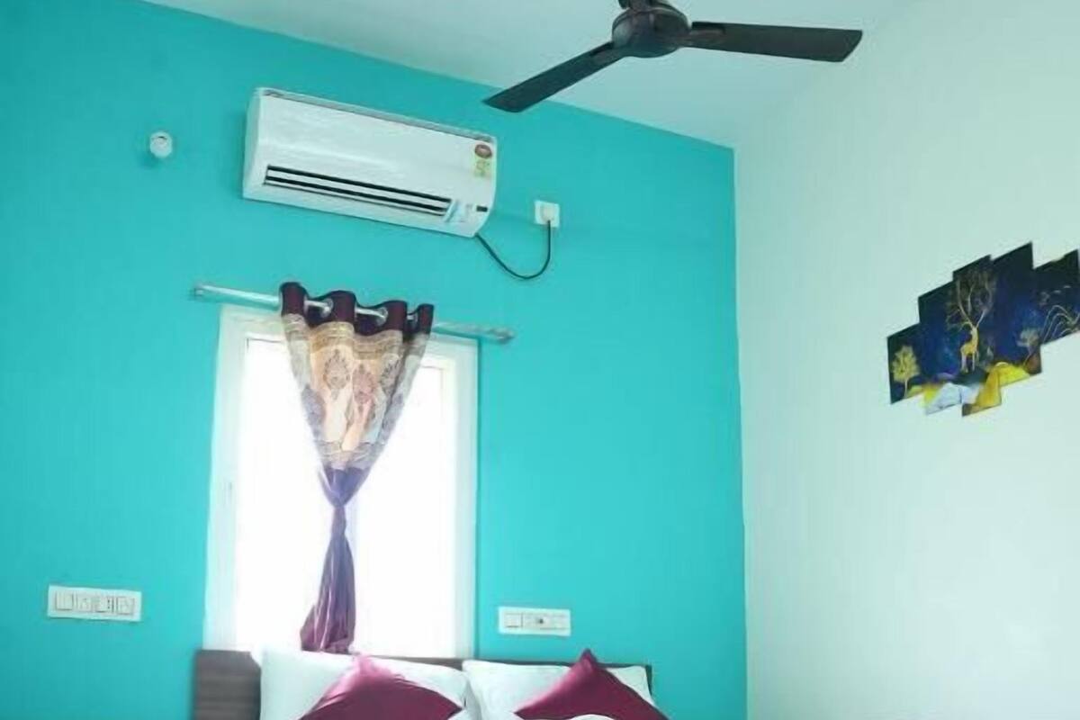 Quarto com cama, ar-condicionado, ventilador de teto, janela com cortinas e um adorno na parede.