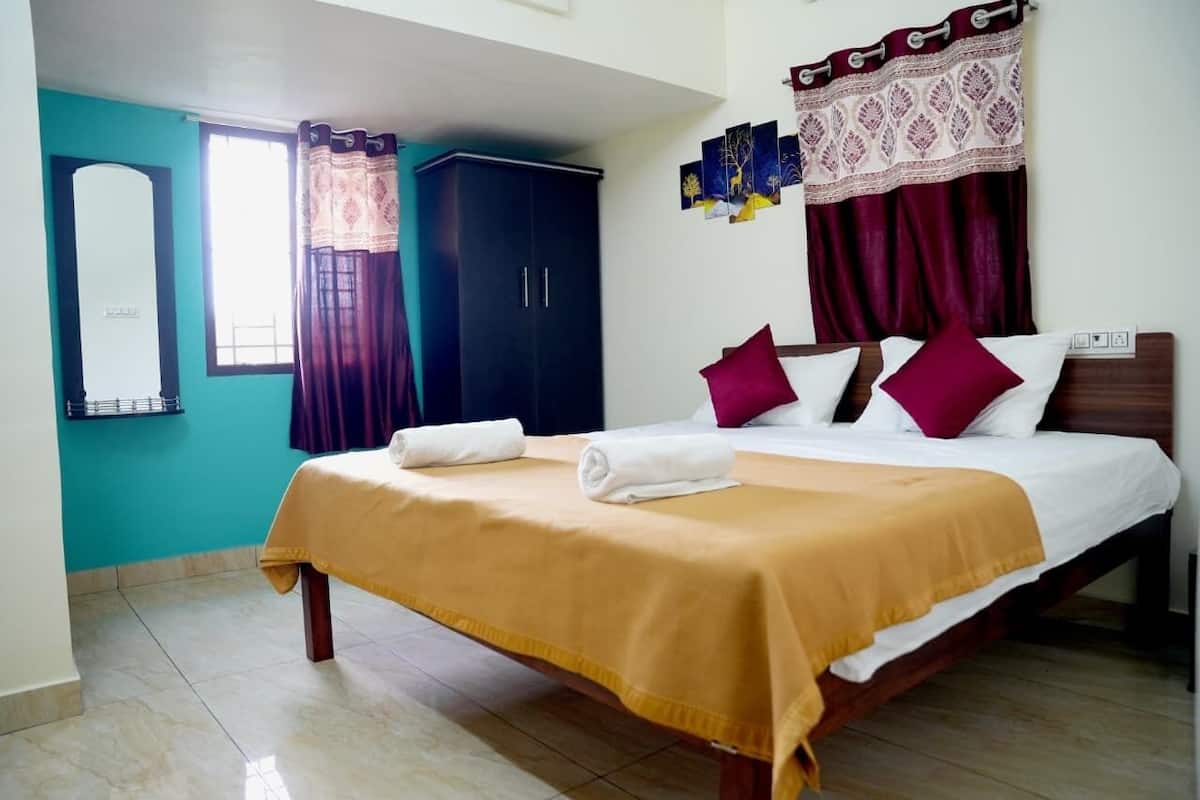 Uma cama bem arrumada com colcha amarela e duas almofadas, um guarda-roupa azul escuro, uma parede verde-água e uma janela com cortinas roxas.