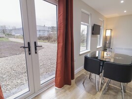 Quarto com porta de vidro que dá para uma área de cascalho, uma mesa com cadeiras, televisão fixada na parede e abajur.