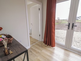 Quarto com piso de madeira, mesa com cesta e vaso, porta de vidro deslizante com cortinas marrons, e porta branca que dá para outro cômodo.