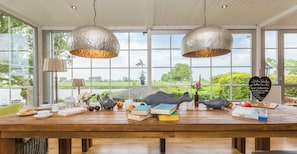 Mesa de madeira com livros, um quebra-cabeça e objetos decorativos em um quarto com grandes janelas e luminárias pendentes.