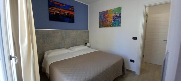 Quarto de hotel com uma cama, mesa de cabeceira, um quadro na parede e uma porta à direita.