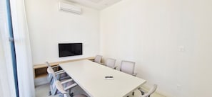 Uma sala de conferências com uma mesa comprida, seis cadeiras, uma TV de tela plana e um ar-condicionado.
