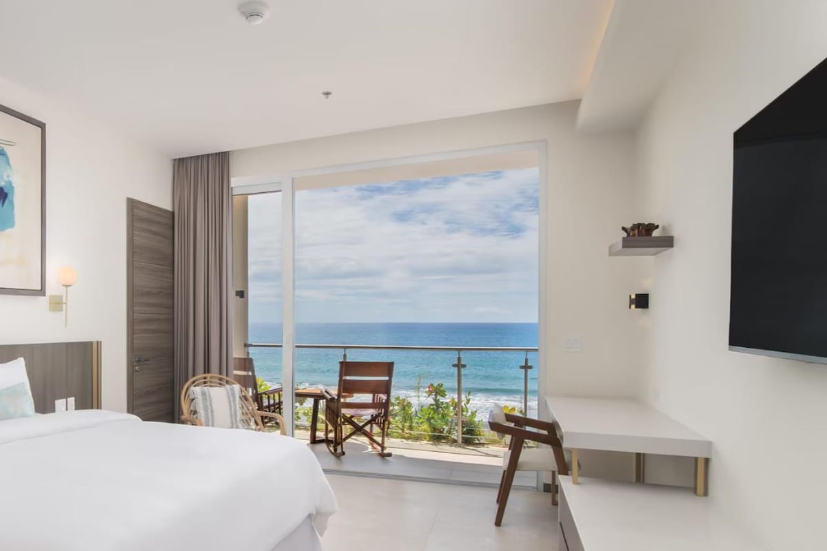 Quarto de hotel com uma cama grande, um quadro, varanda com vista para o oceano e um espaço para relaxar.