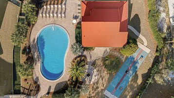 Vista aérea de uma piscina com um terraço ao redor, um edifício com telhado vermelho e uma quadra de tênis.